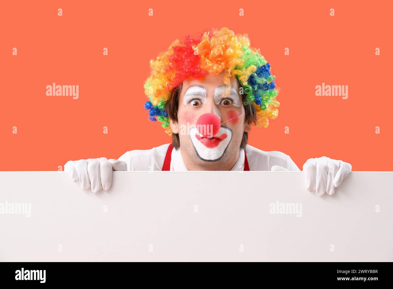 Portrait of surprised clown with blank poster on orange background ...