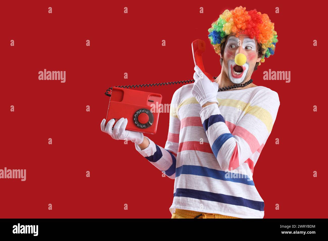 Portrait of surprised clown with retro telephone on red background ...