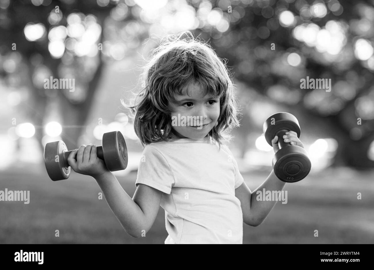 Cute little boy doing exercises with dumbbells in green park. Closeup ...
