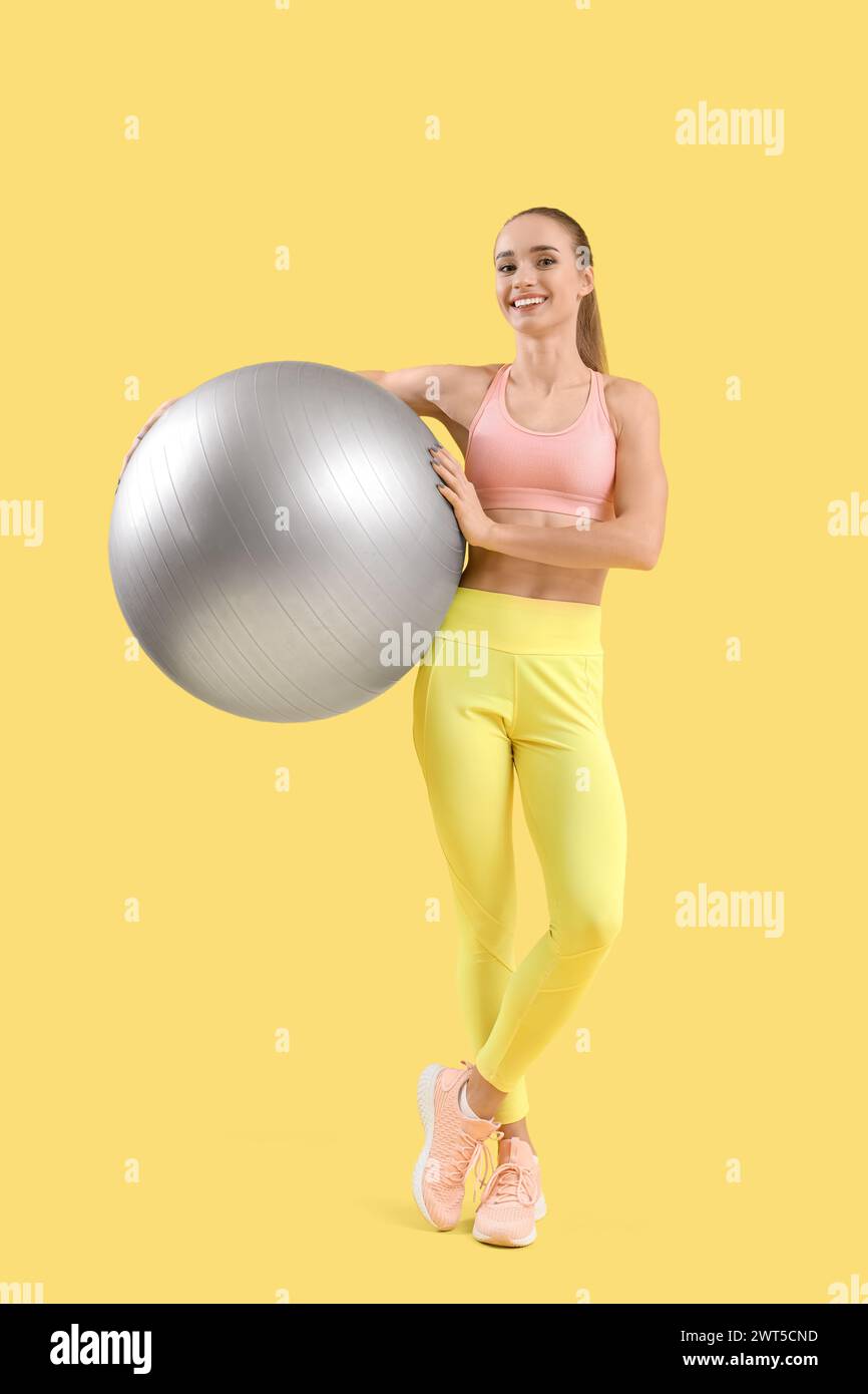 Portrait of female fitness trainer with fitball on yellow background ...