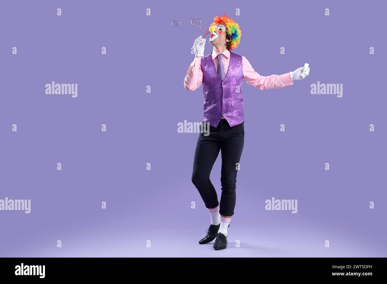 Portrait of clown blowing soap bubbles on lilac background. April Fool ...