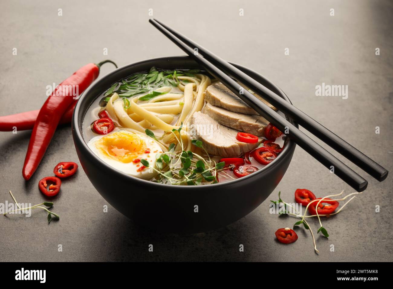 Delicious ramen with meat in bowl, chopsticks and chili peppers on grey textured table. Noodle ...