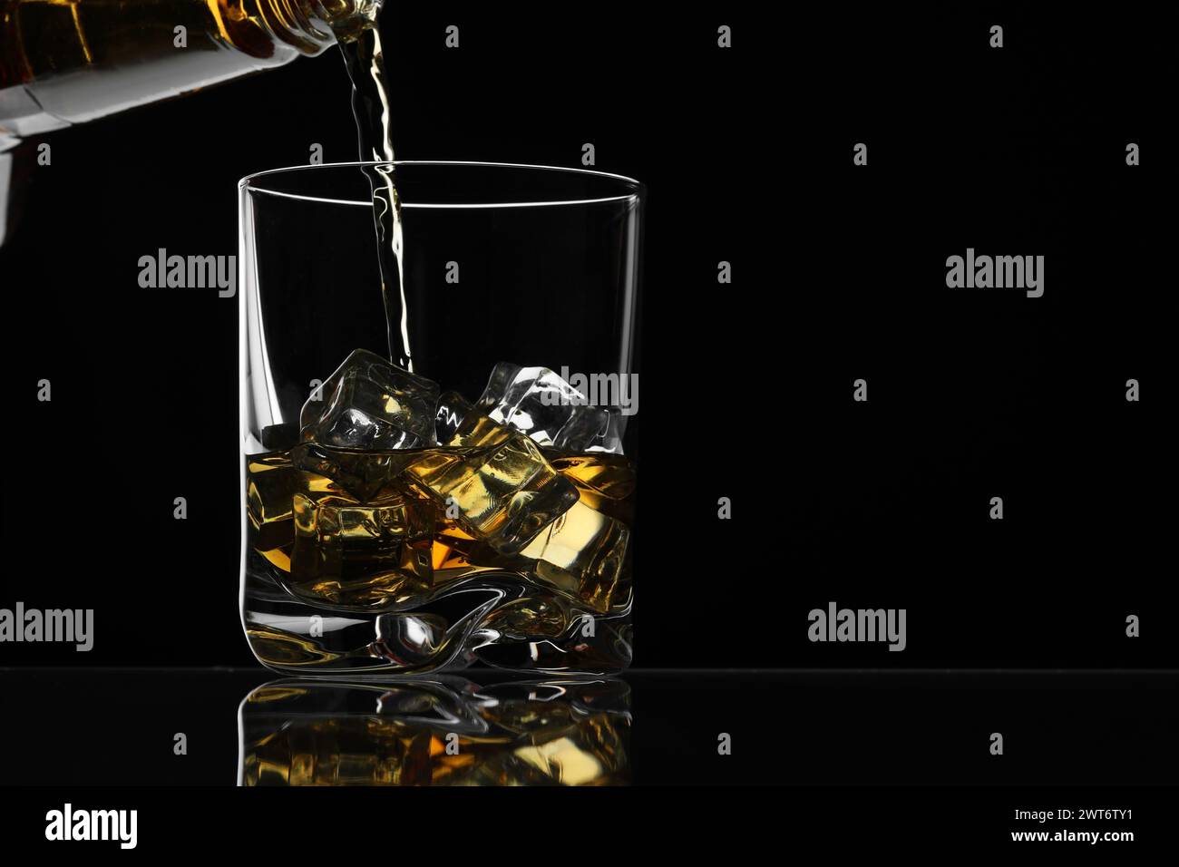 Pouring whiskey into glass with ice cubes at table against black background, space for text ...