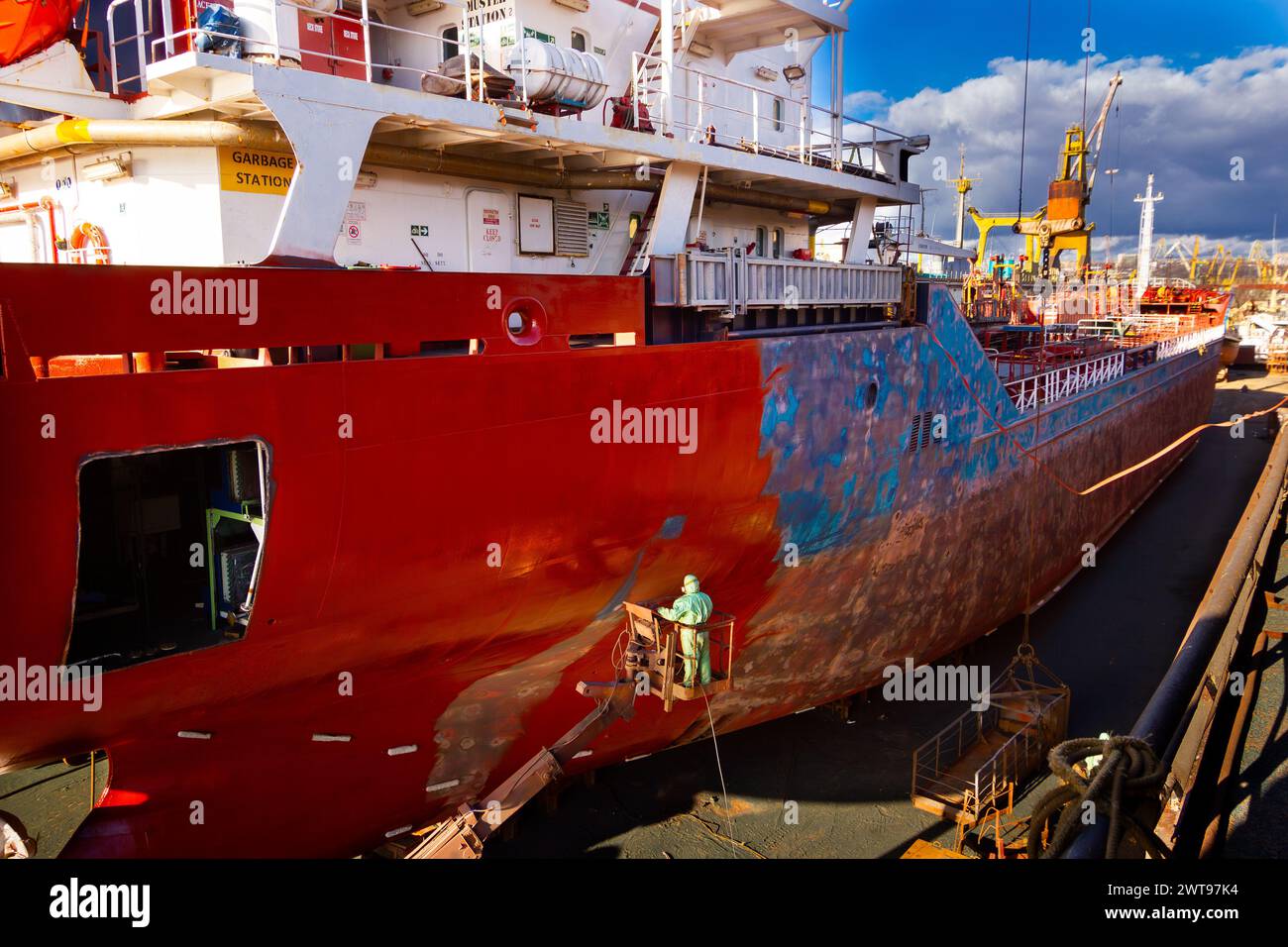 Worker paints cargo ship hull red in dry dock under blue sky. Maritime ...