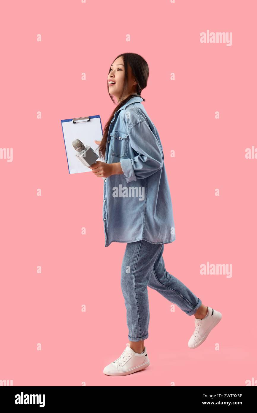 Female Asian journalist with microphone and clipboard walking on pink ...