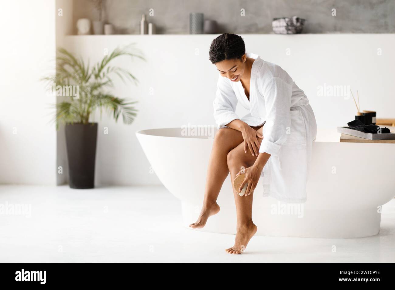 Smiling black woman in bathrobe dry brushing legs, practicing skin ...