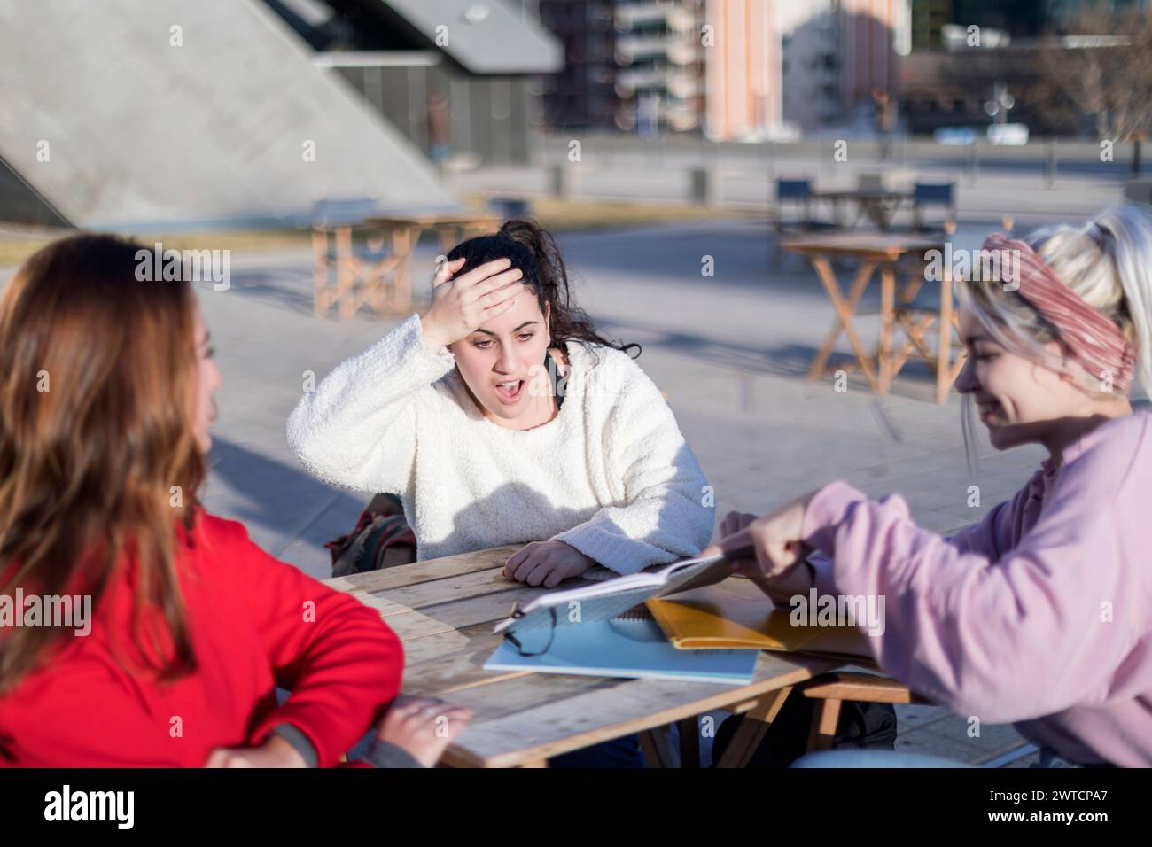 Three women sit at an outdoor table; one in white looks surprised while ...