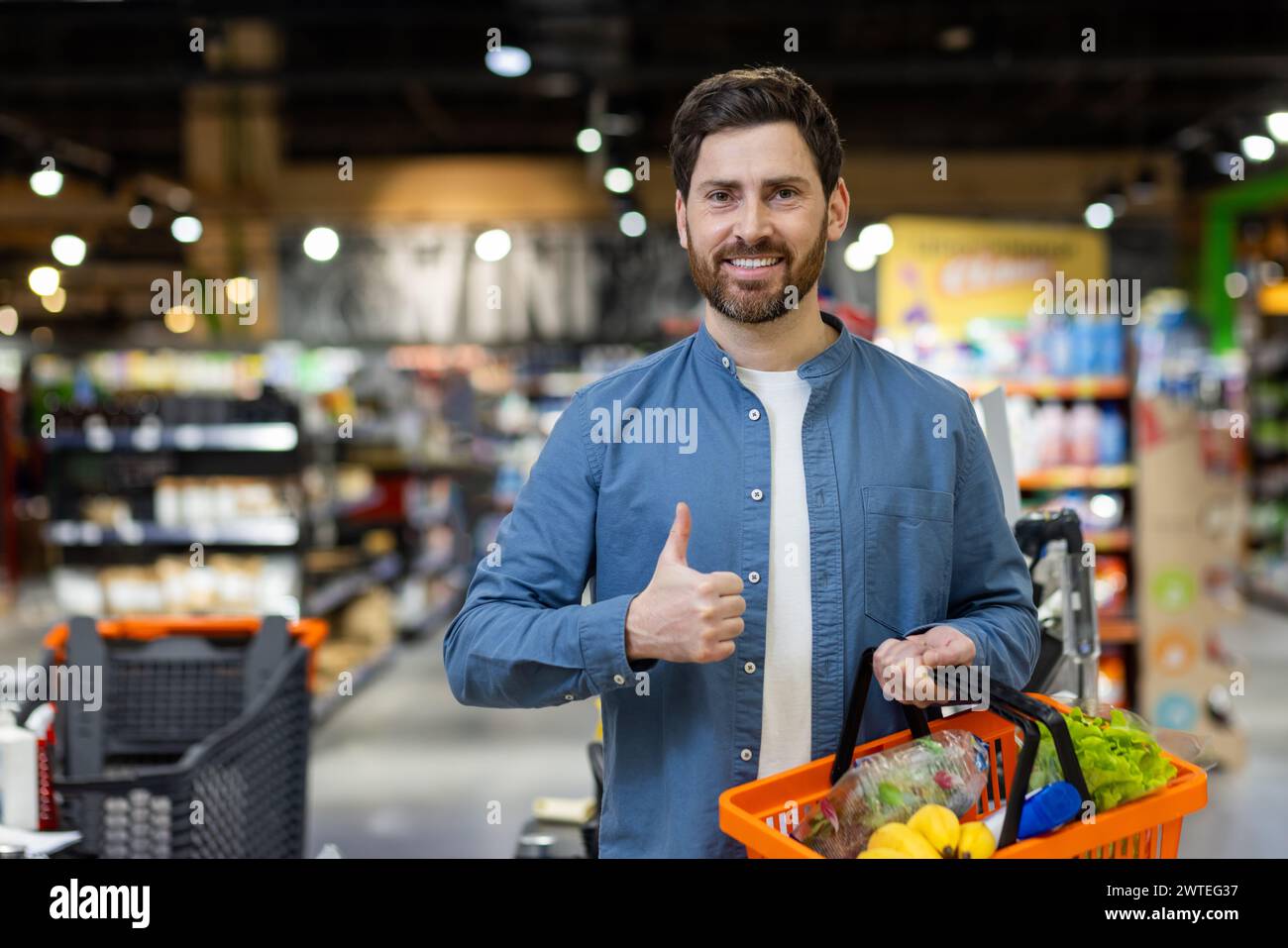 A man in a supermarket with a full shopping basket gives a thumbs up ...