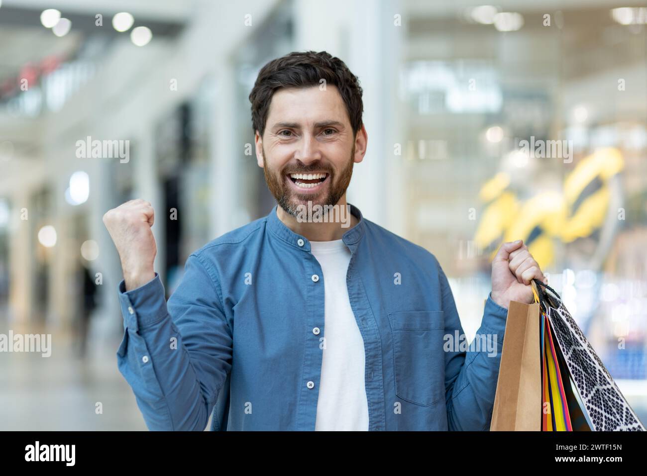 Cheerful bearded man in a mall holding colorful shopping bags, feeling ...