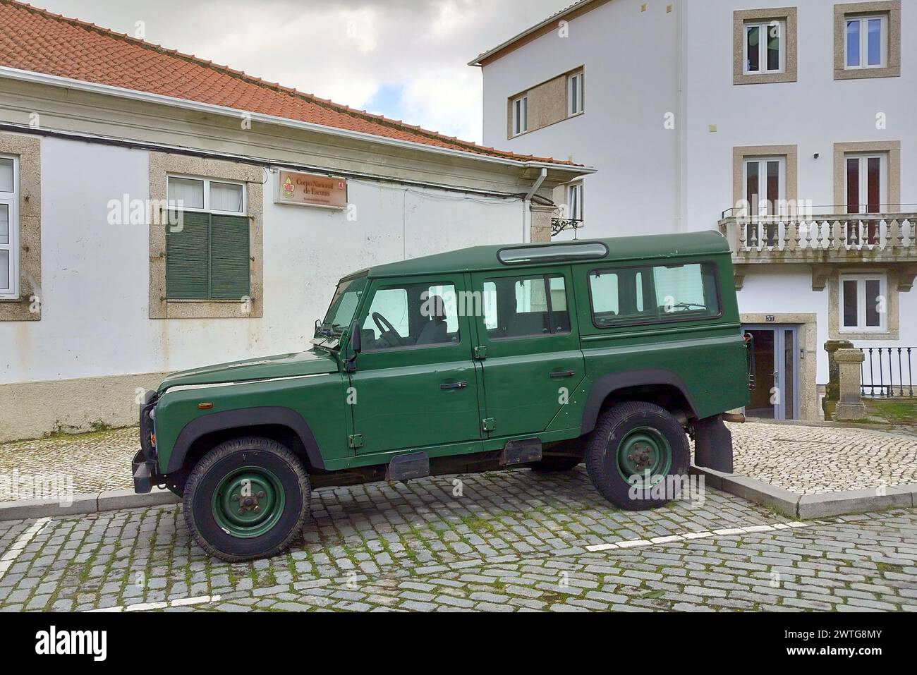Green rugged Land Rover Defender parked by a sidewalk on a sloping ...