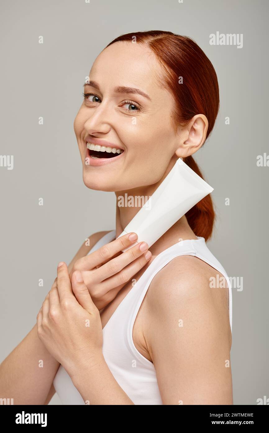 excited redhead woman in 30s holding tube with body lotion on grey ...