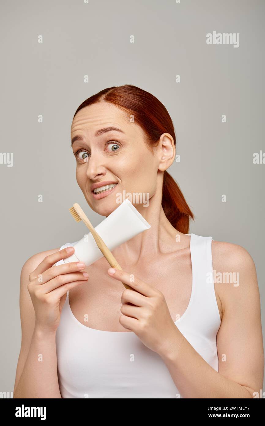 confused redhead woman in tank top holding toothpaste and toothbrush ...