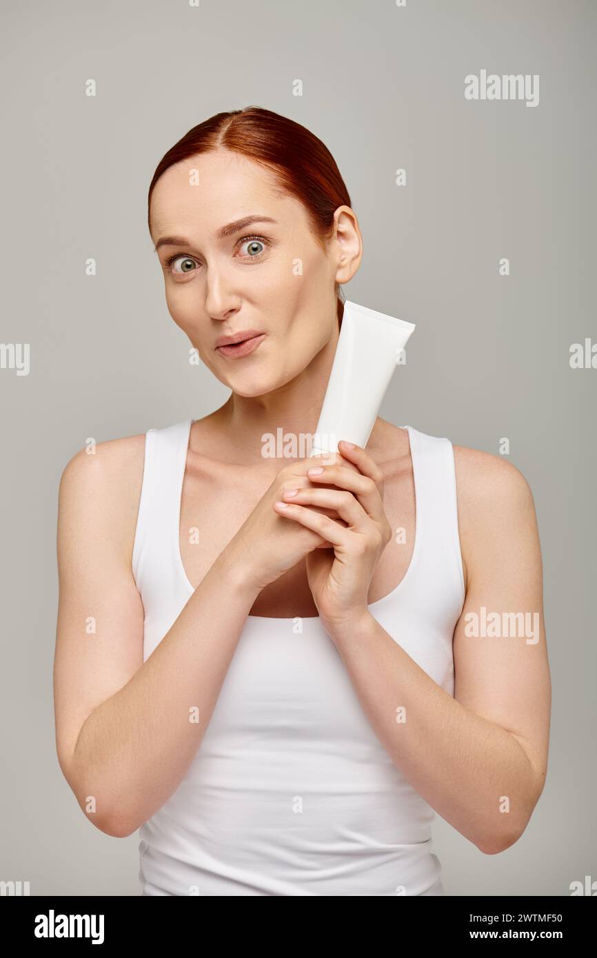 amazed young woman with red hair showing tube with body lotion on grey ...