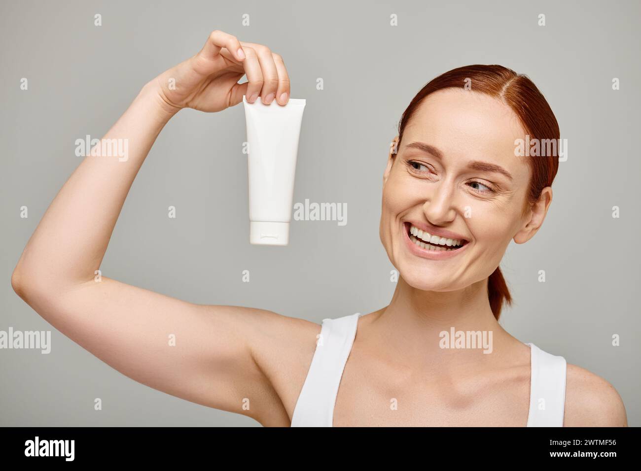 Redhead and cheerful woman in 30s showing tube with body lotion on grey ...