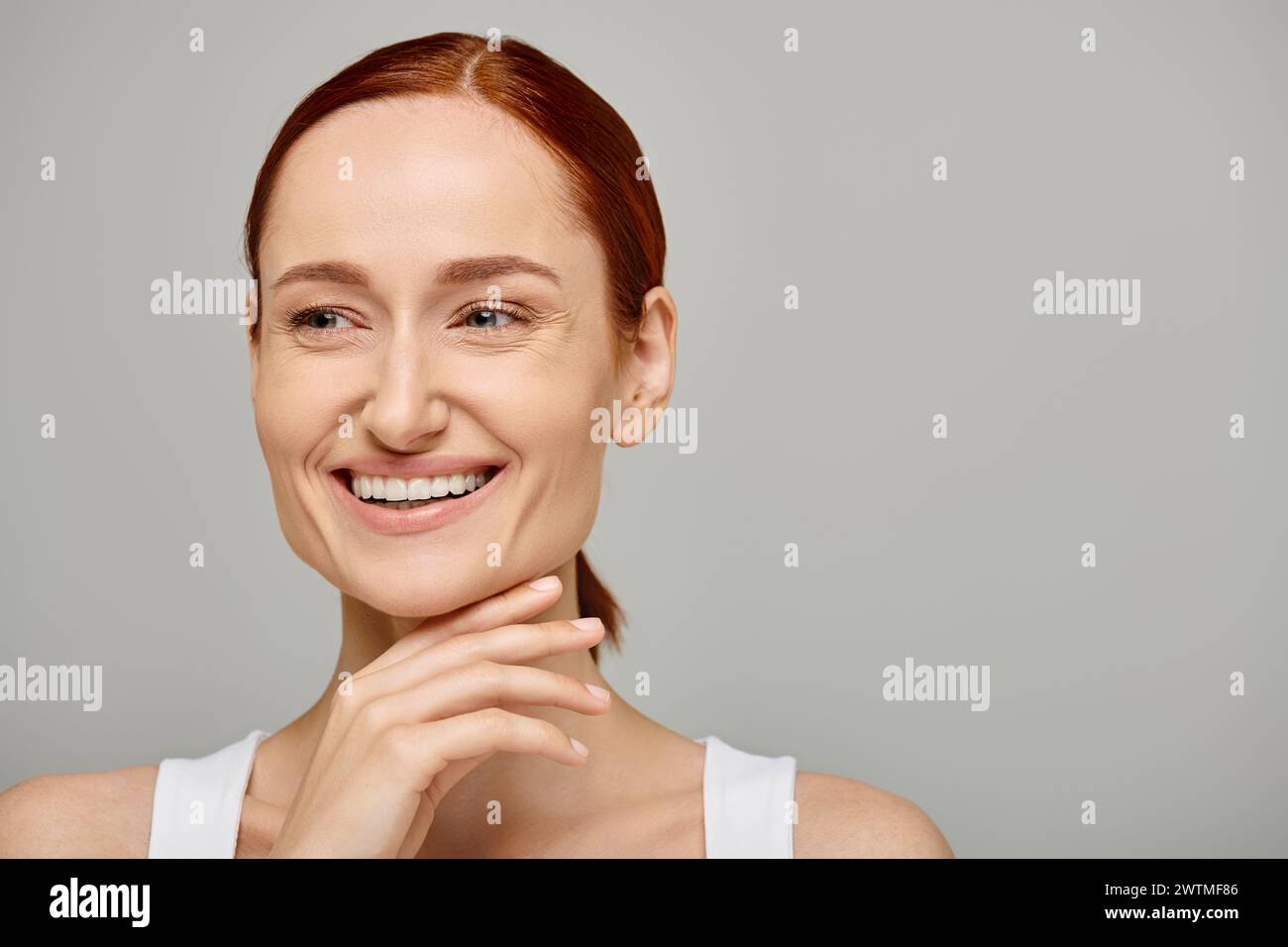 cheerful and redhead woman in her 30s posing in white tank top on grey ...