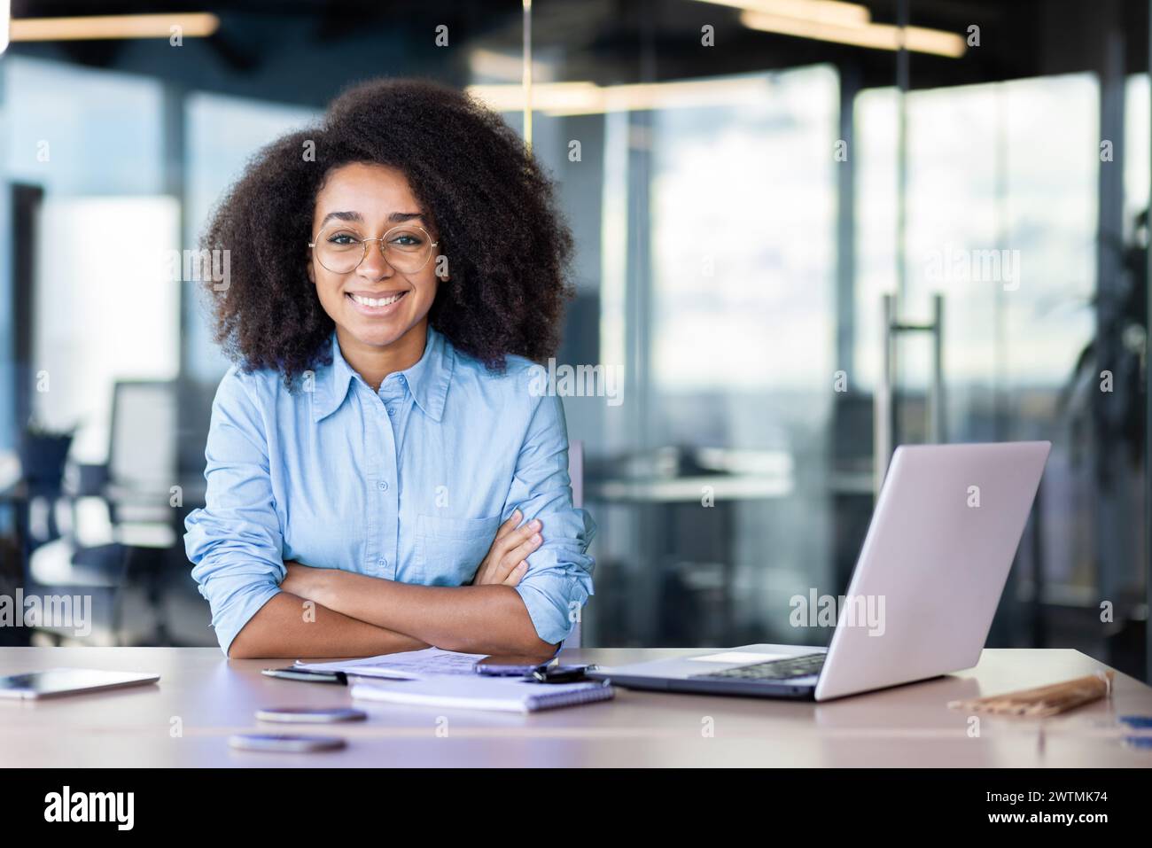 Successful black woman in blue shirt leaning with folded hands on table ...