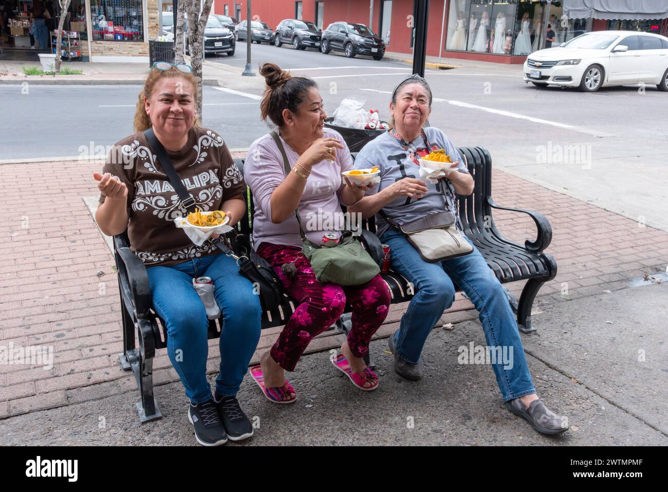 Three smiling middle-aged women eating nachos, sitting on a metal bench ...