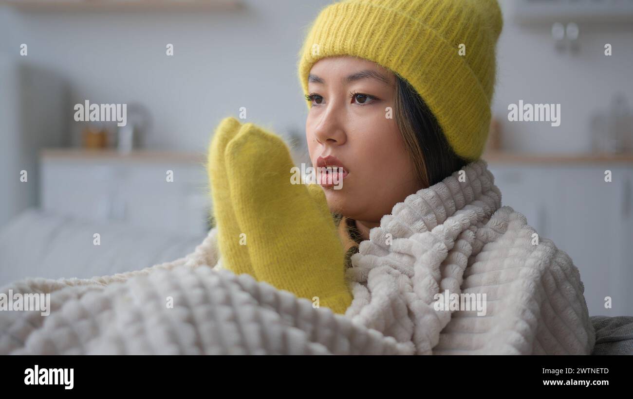 Sick Asian woman in warm hat and mittens freezing sit in cold kitchen ...