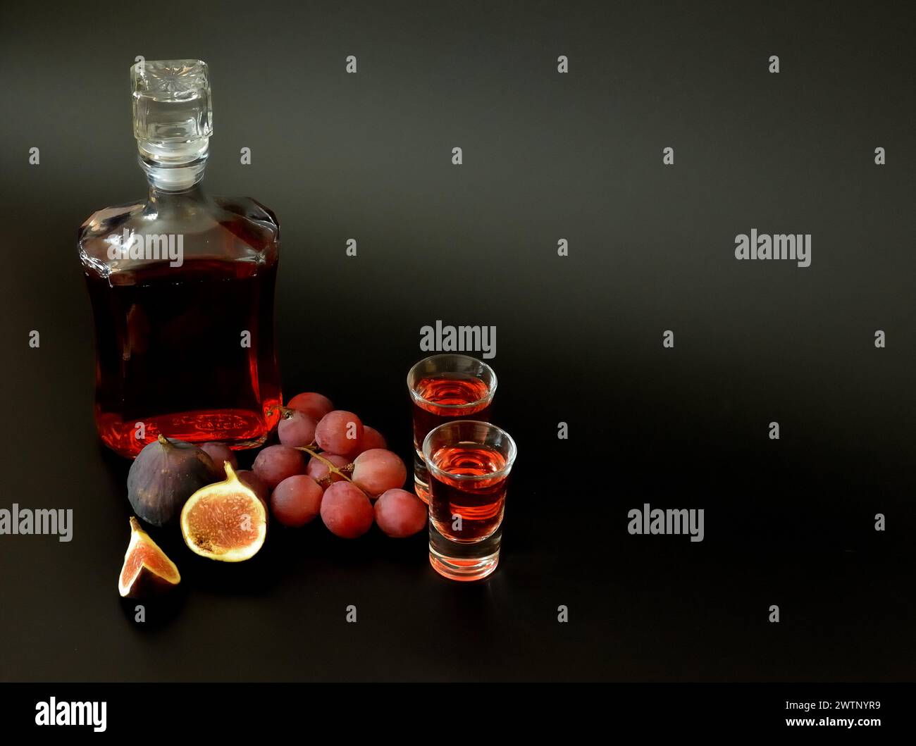 Two glasses and a glass decanter with homemade liqueur on a black background, next to pieces of ...