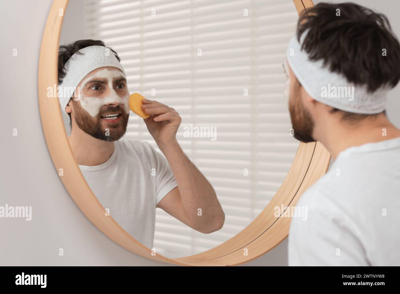 Man with headband washing his face using sponge near mirror in bathroom ...