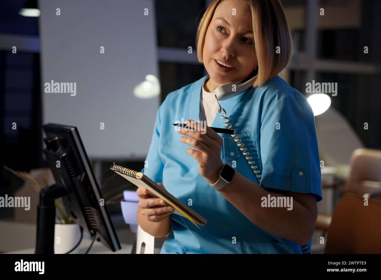 Female nurse with notebook talking by telephone at hospital in evening ...