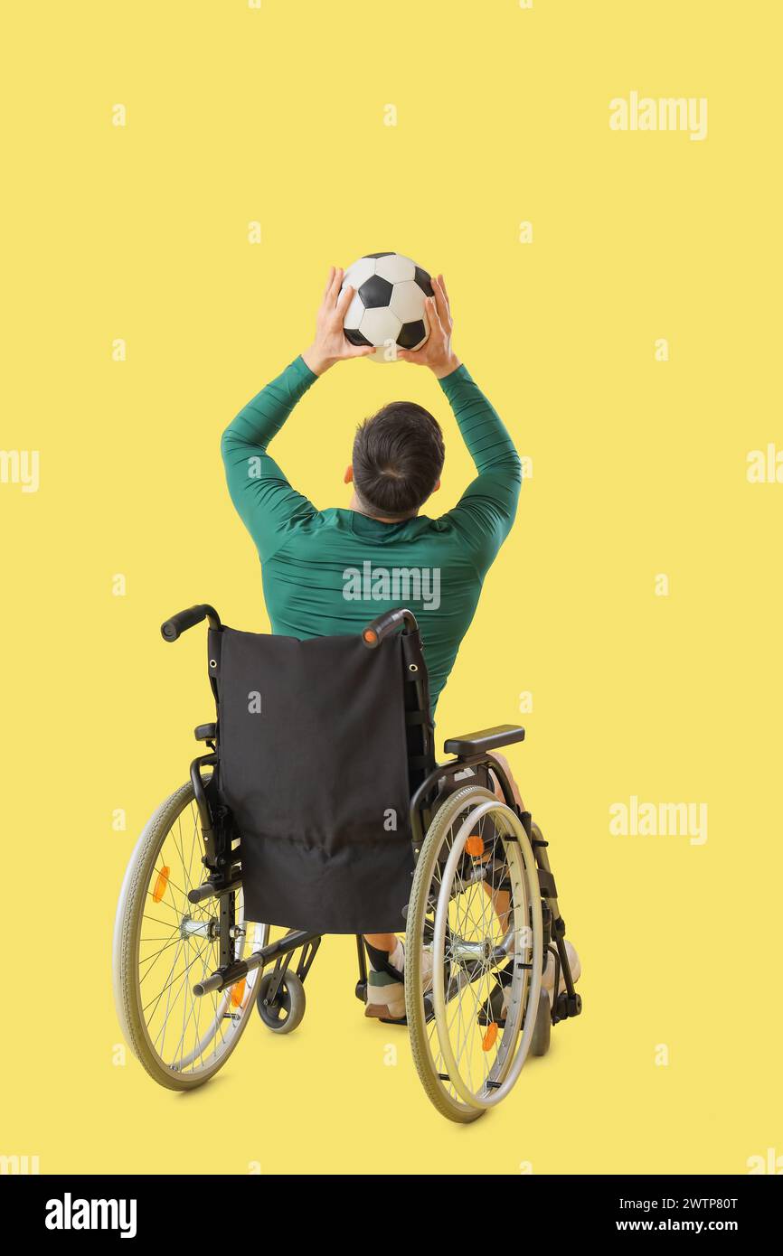 Male footballer in wheelchair with ball on yellow background, back view ...