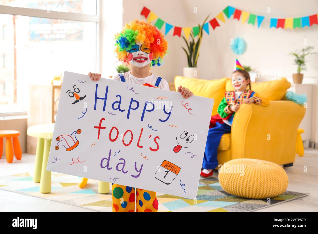 Funny little boy in clown costume with poster at home. April Fools' Day ...