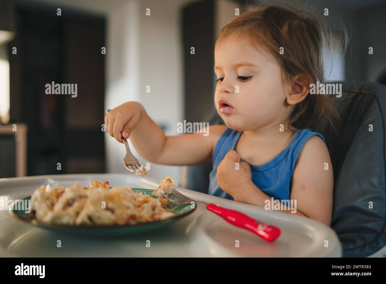 Young girl enjoying pasta in high chair with fork, Tableware Stock ...