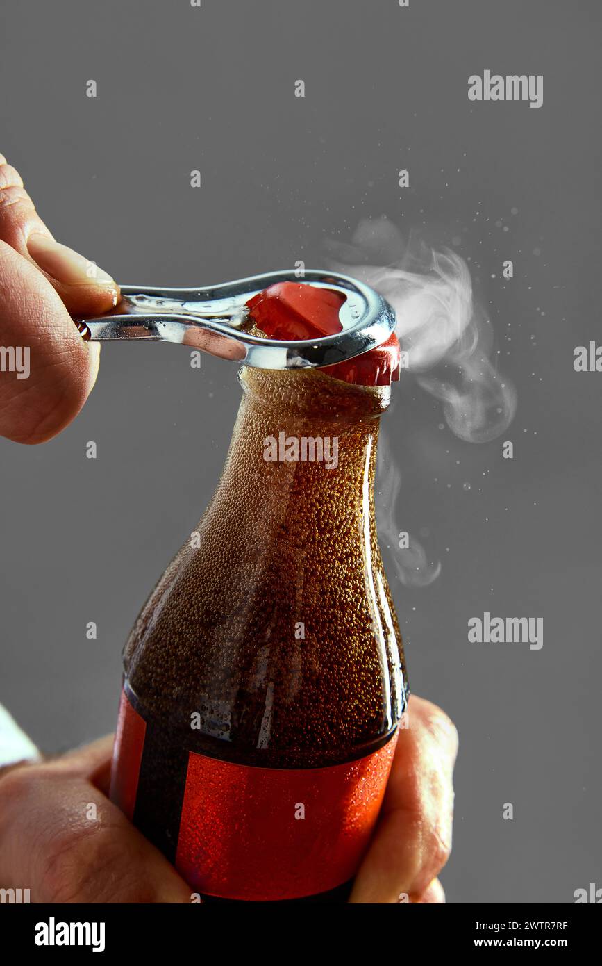 Hand opening bottle of soda, coke, beer with bottle opener against gray ...