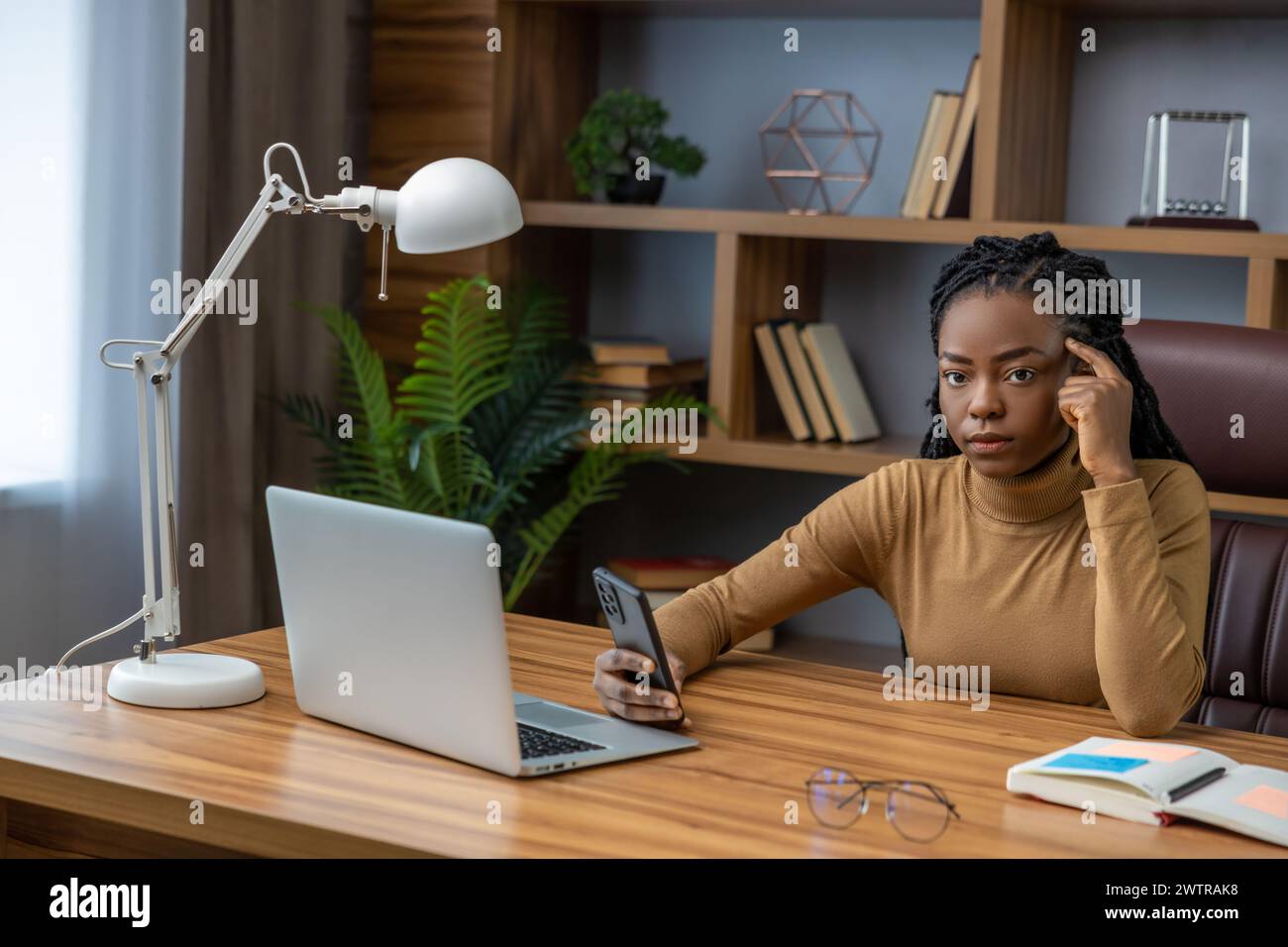 Pensive woman with dreadlocks sitting at workplace with laptop and ...