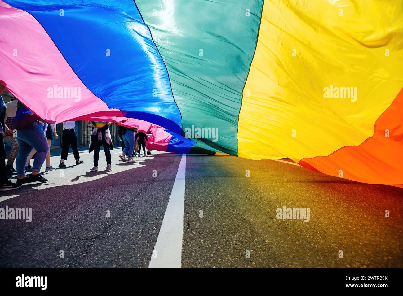 The sun filters through a large rainbow pride flag held aloft by people ...