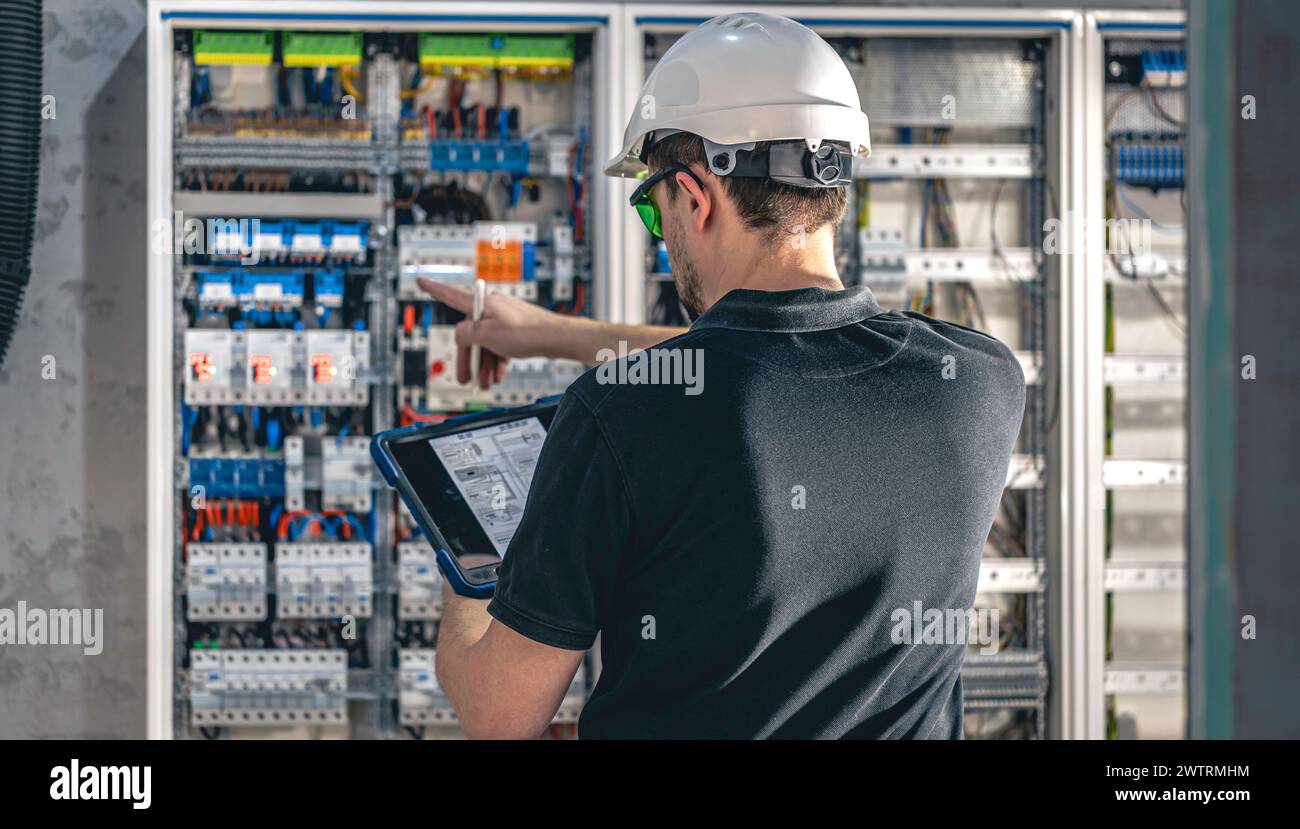 Man, an electrical working in a switchboard with fuses, uses a tablet ...