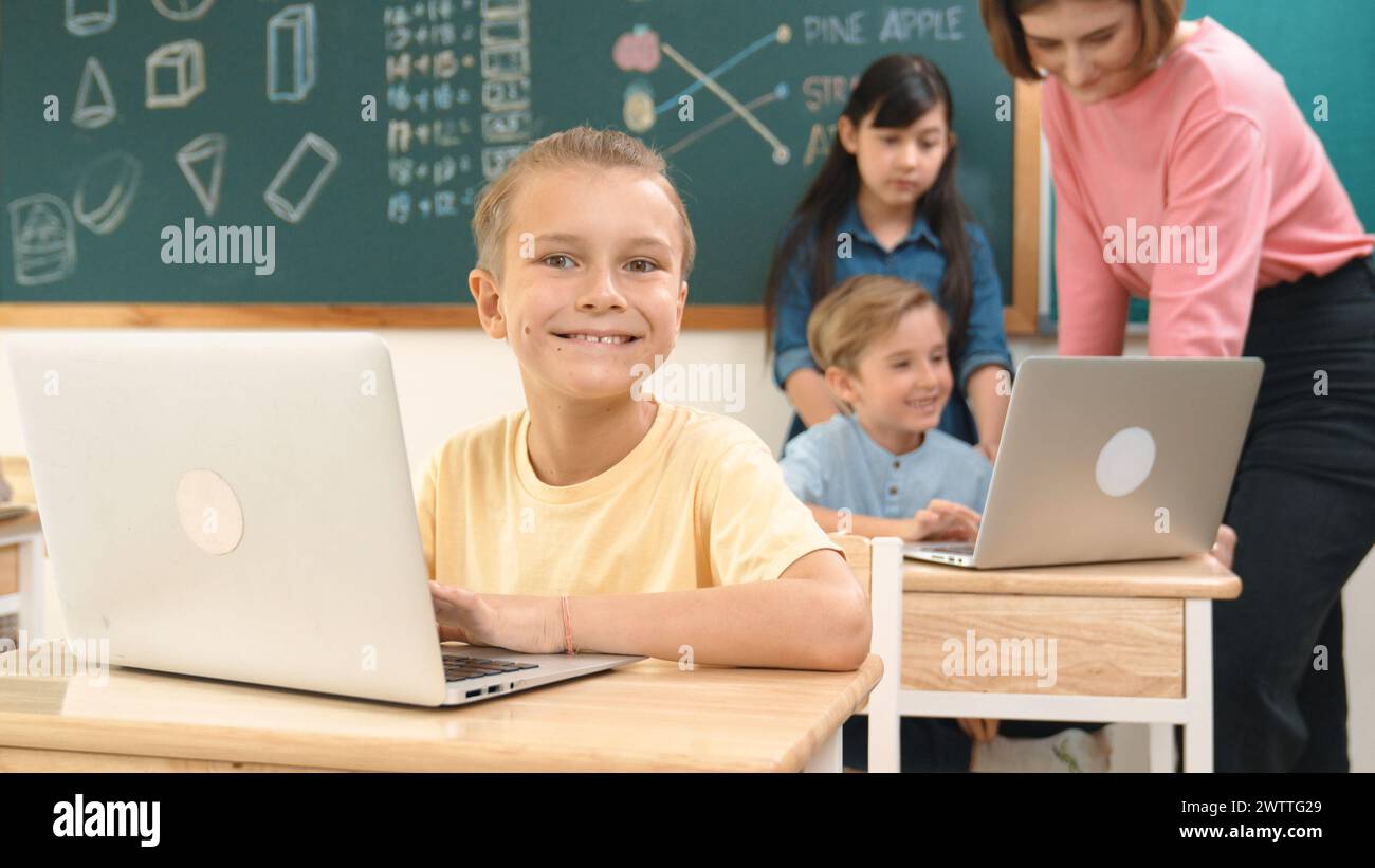 Caucasian boy focus on programing system while looking at camera surrounded by diverse student ...