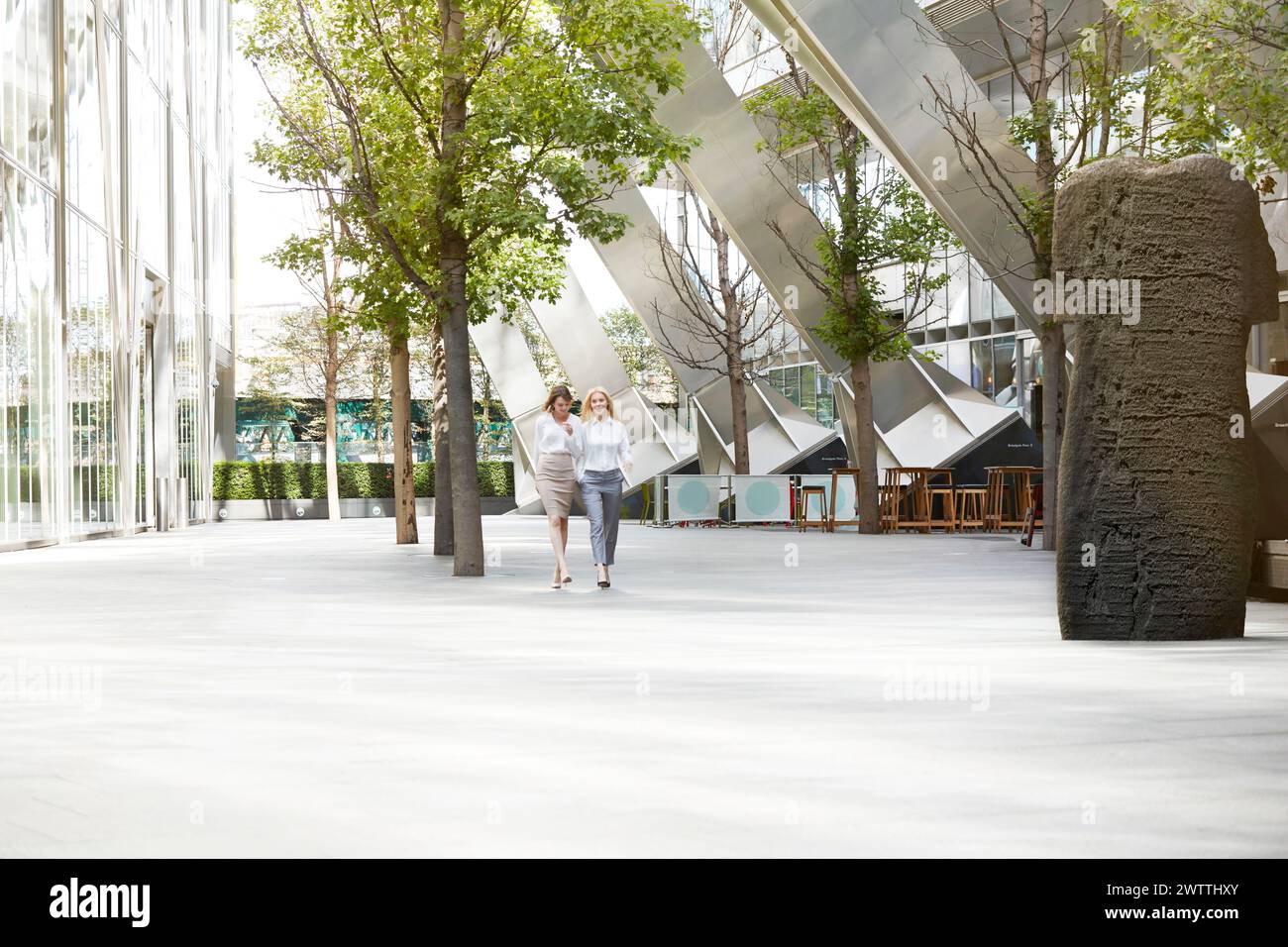 Two people walking in a modern plaza with trees and unique architecture ...