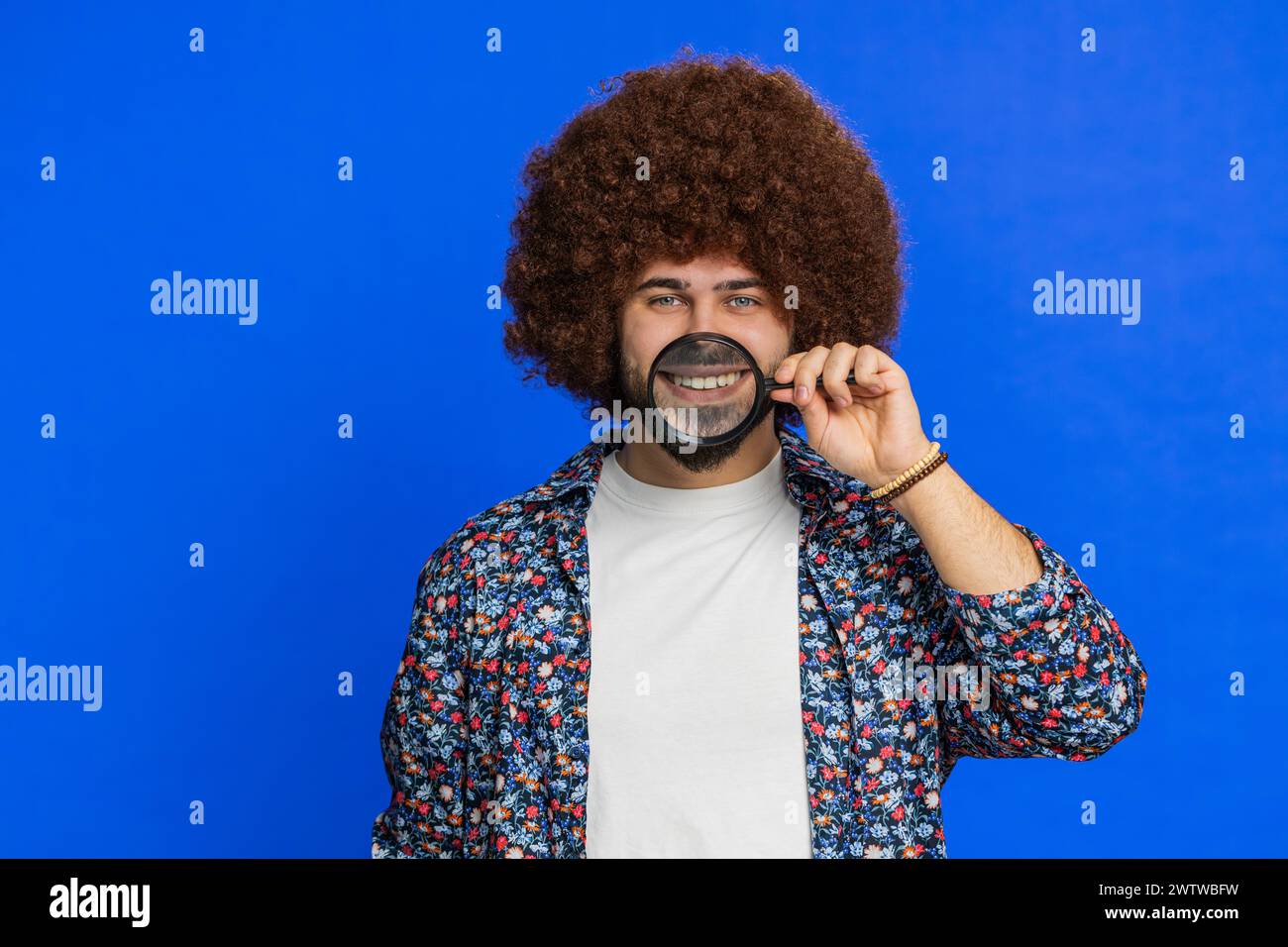 Happy man with Afro hairstyle wig holding magnifier glass on teeth ...