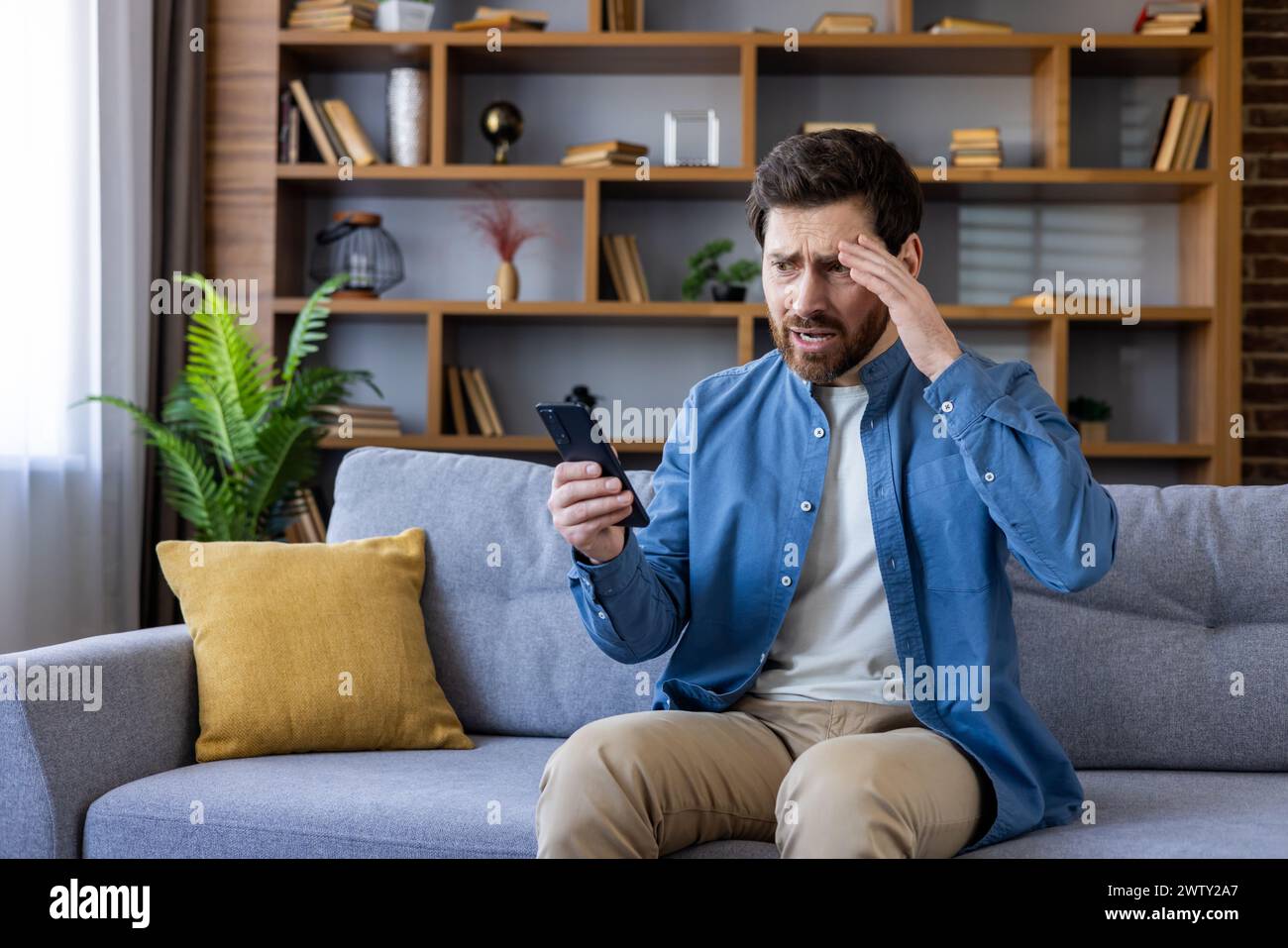 Stressed male sitting on a gray couch at home, expressing anxiety while ...