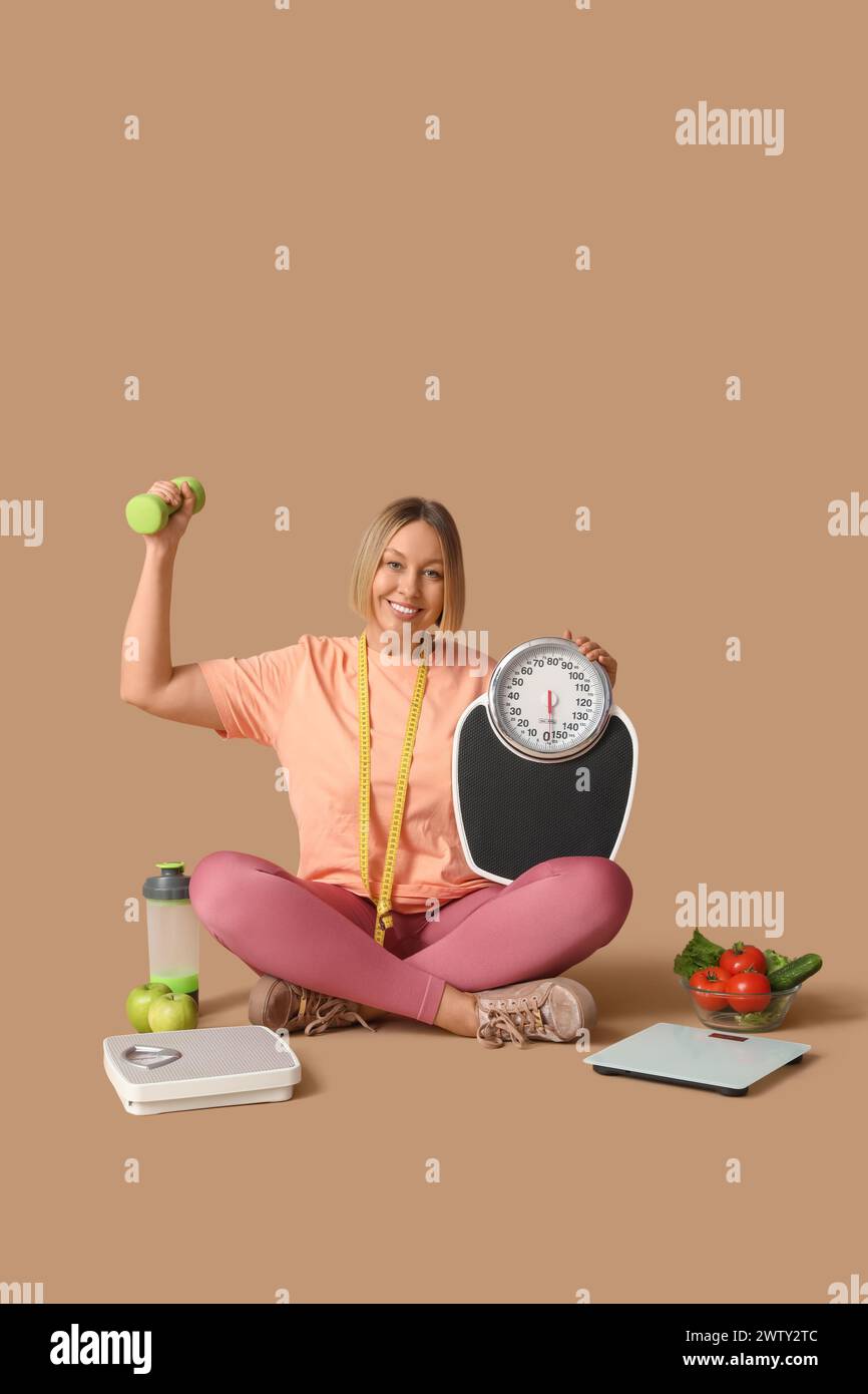 Pretty young woman with different scales and dumbbells on beige ...
