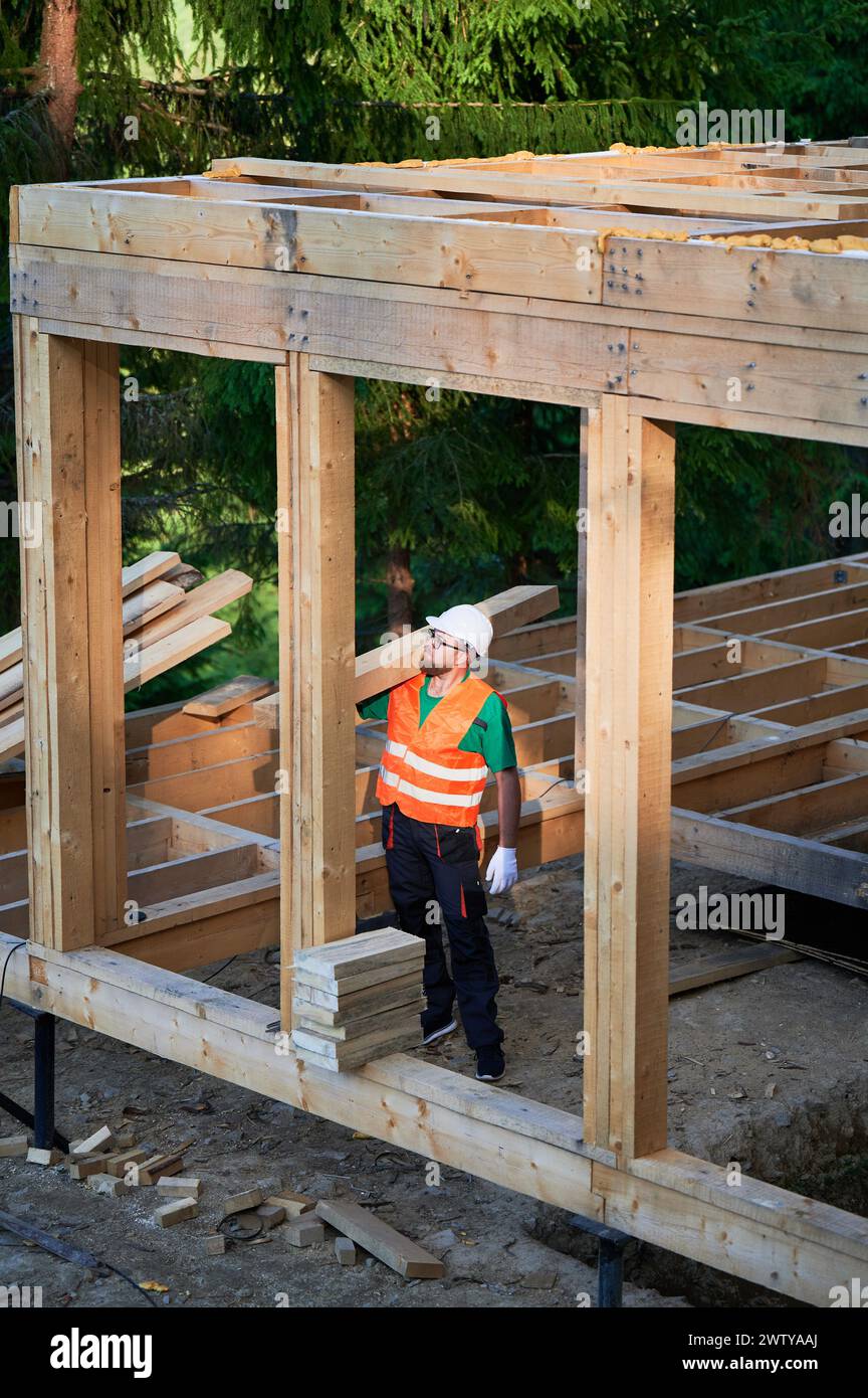Carpenter builds wooden frame house near the forest. Man holds large ...