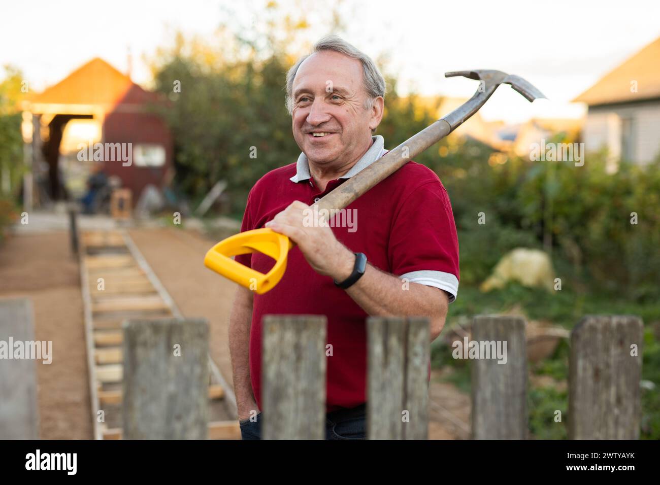 Portrait of satisfied elderly farmer with shovel near wooden fence of ...