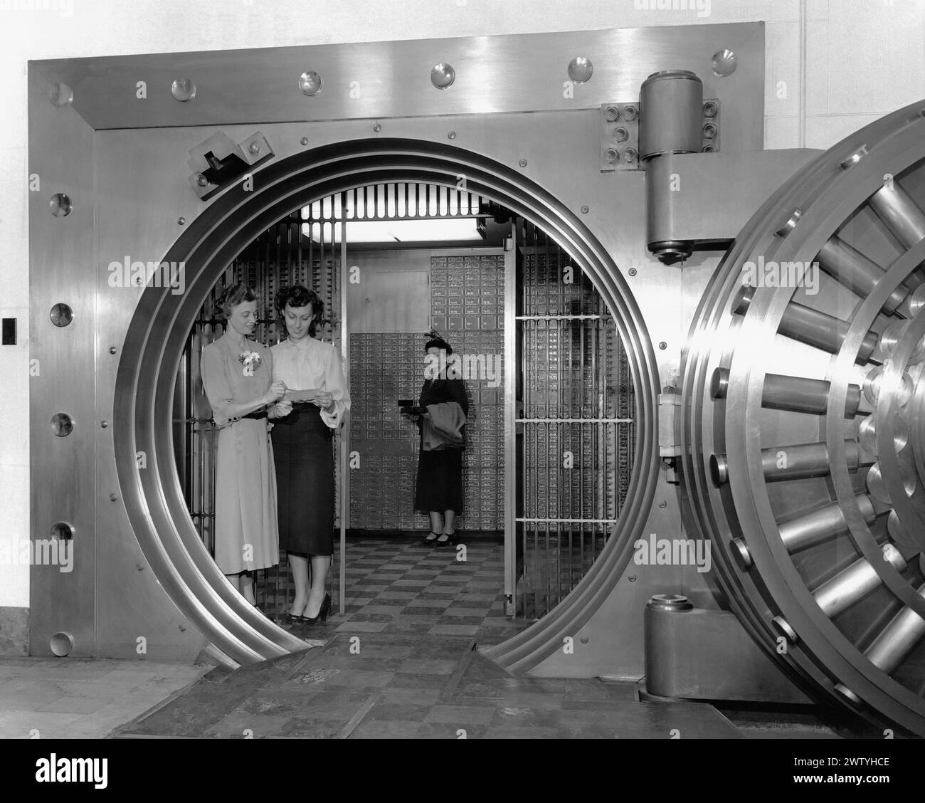 Two young women standing just inside a large bank vault. An older women ...
