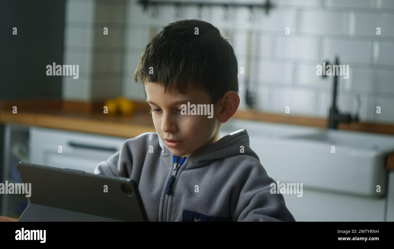 Cute kid sit in the kitchen playing mobile game on digital tablet ...