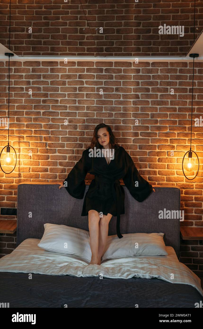 A woman is seated on a bed placed against a brick wall, creating a ...