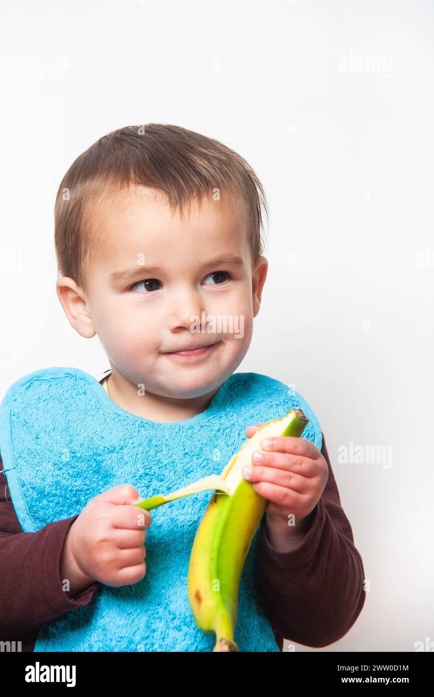Vertical photograph of a boy with a blue bib peeling a banana to eat it ...