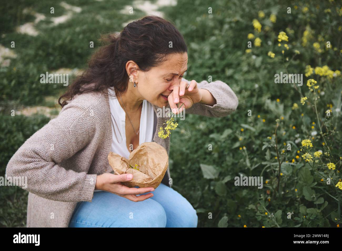 Young woman sneezing while gathering wildflowers in the meadow outdoors, suffering from allergy ...