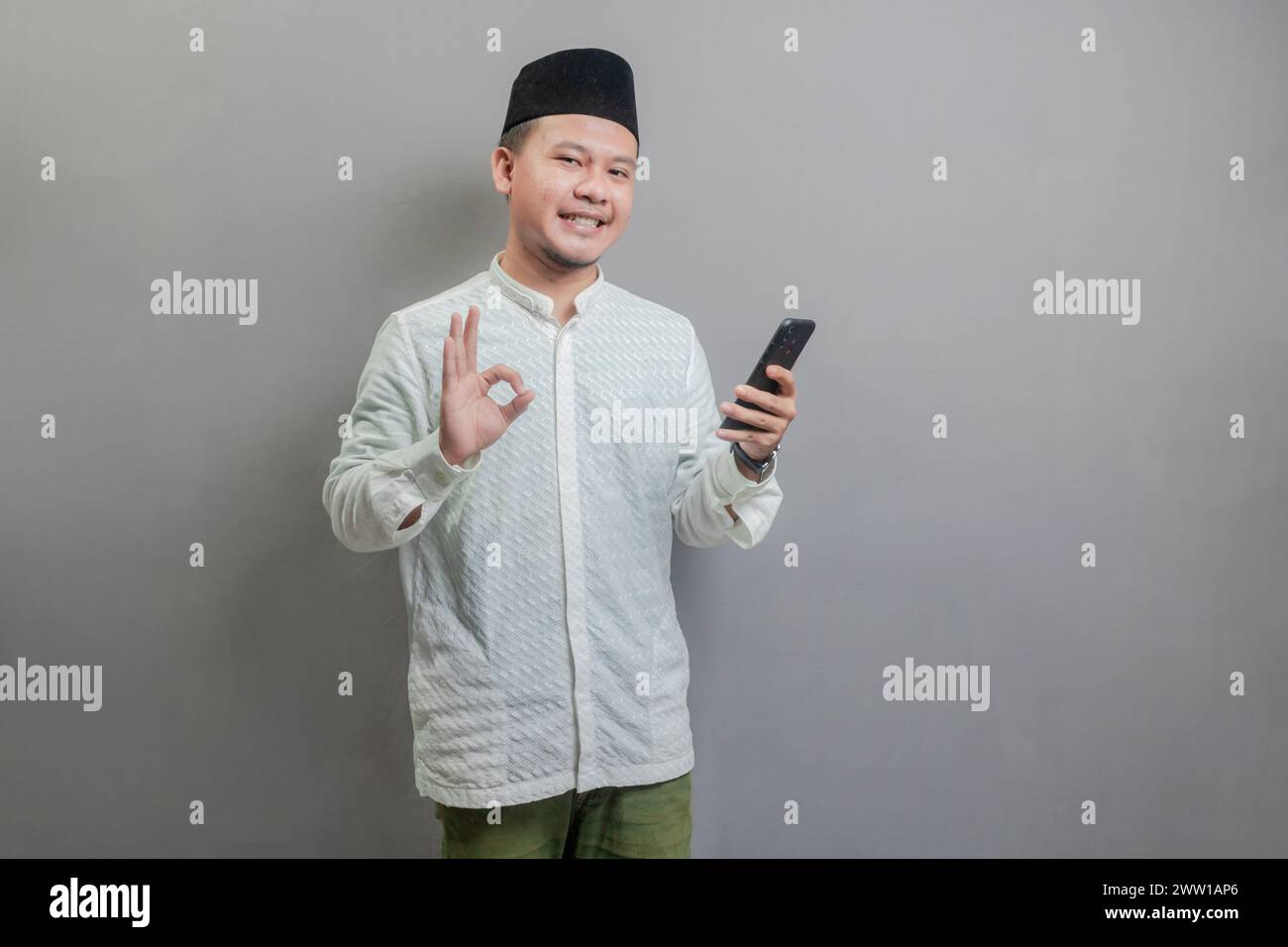 Asian Muslim man standing with ok sign and holding smartphone, wearing a koko shirt and peci ...