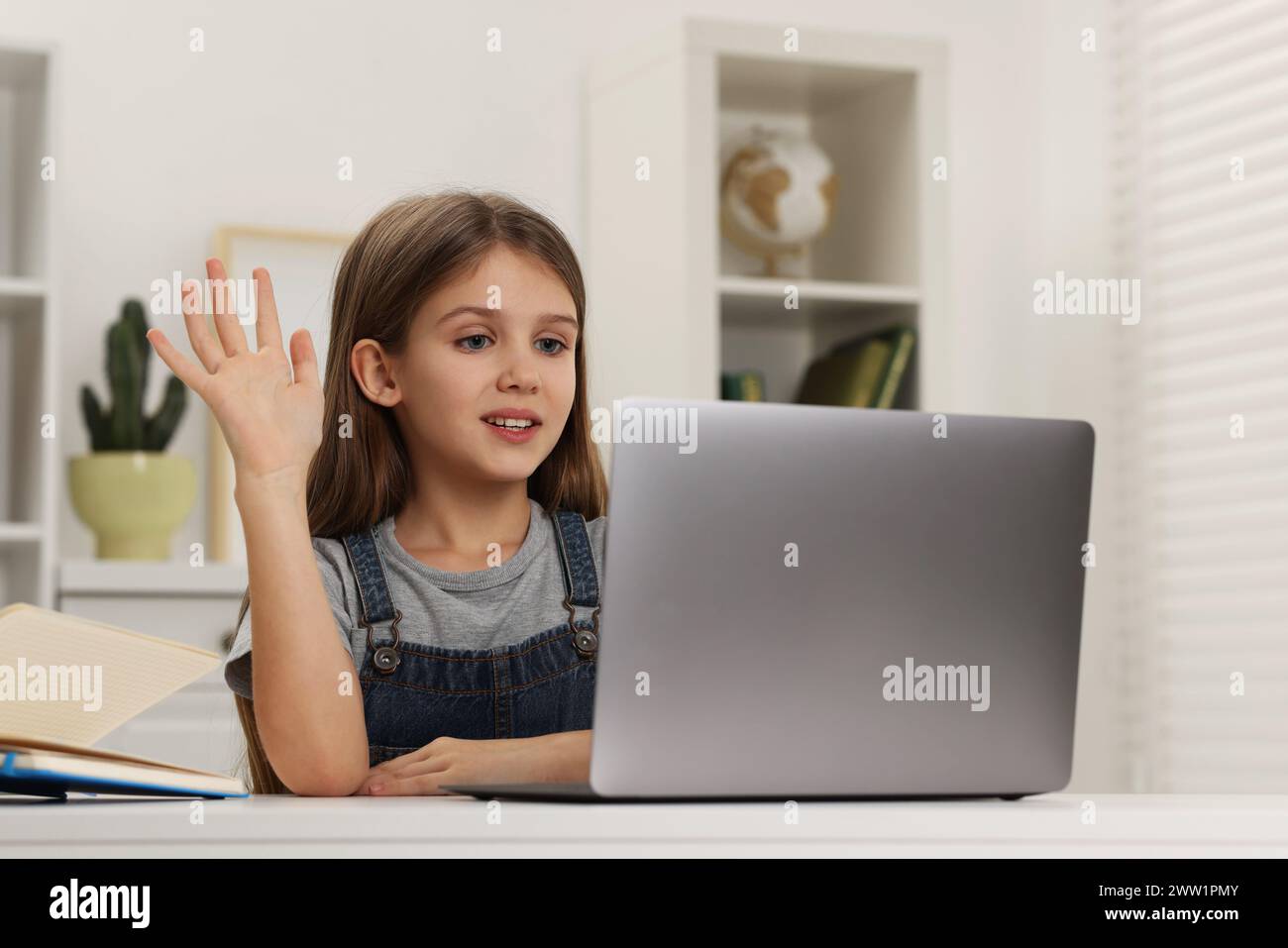 Cute girl waving hello during online lesson via laptop at white table ...