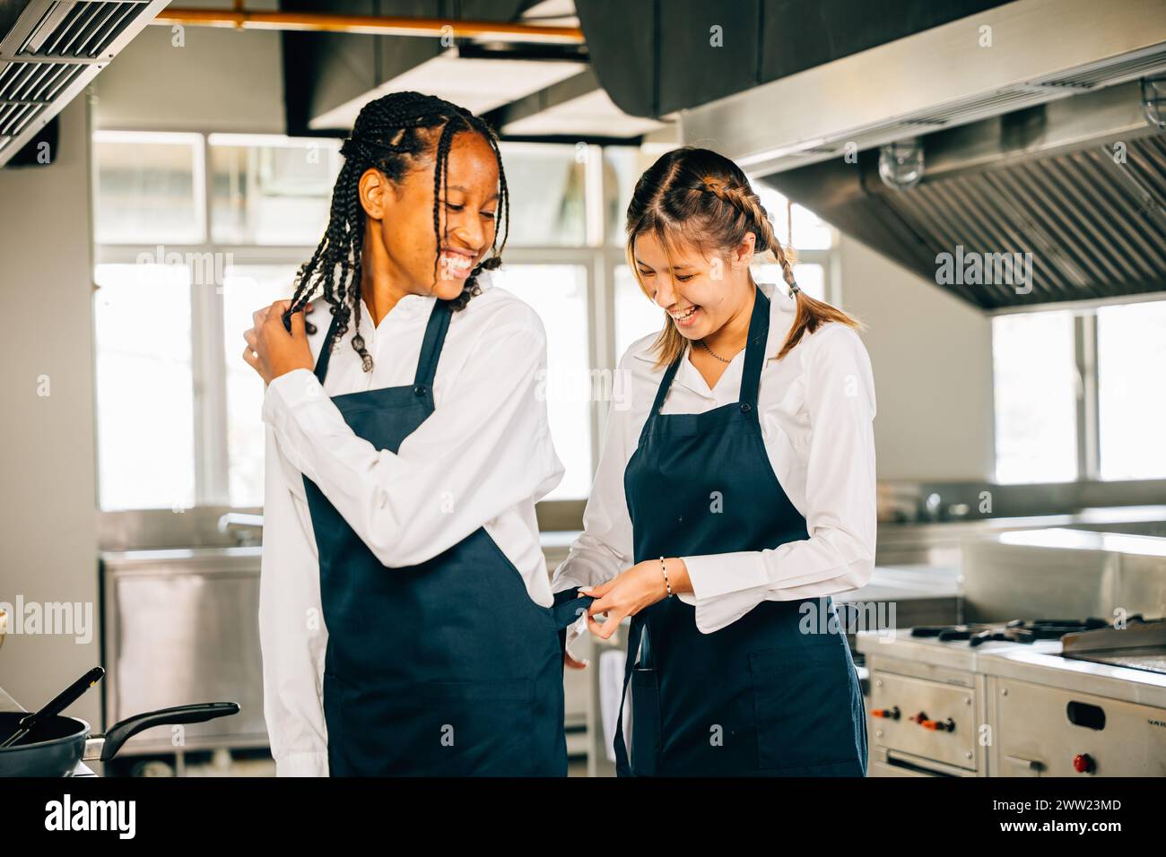 Female chef helps friend tie apron in a restaurant kitchen. Two adults ...