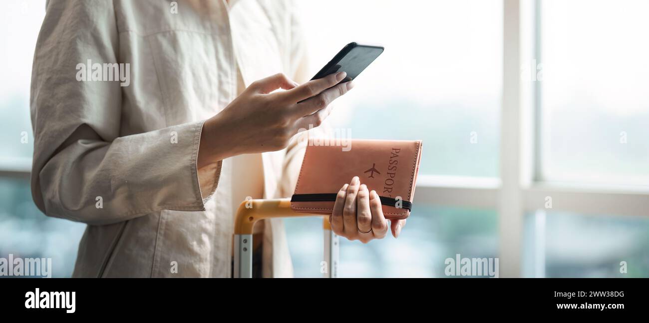 Tourist woman using mobile smartphone and holding passport with suitcase traveling between waits ...