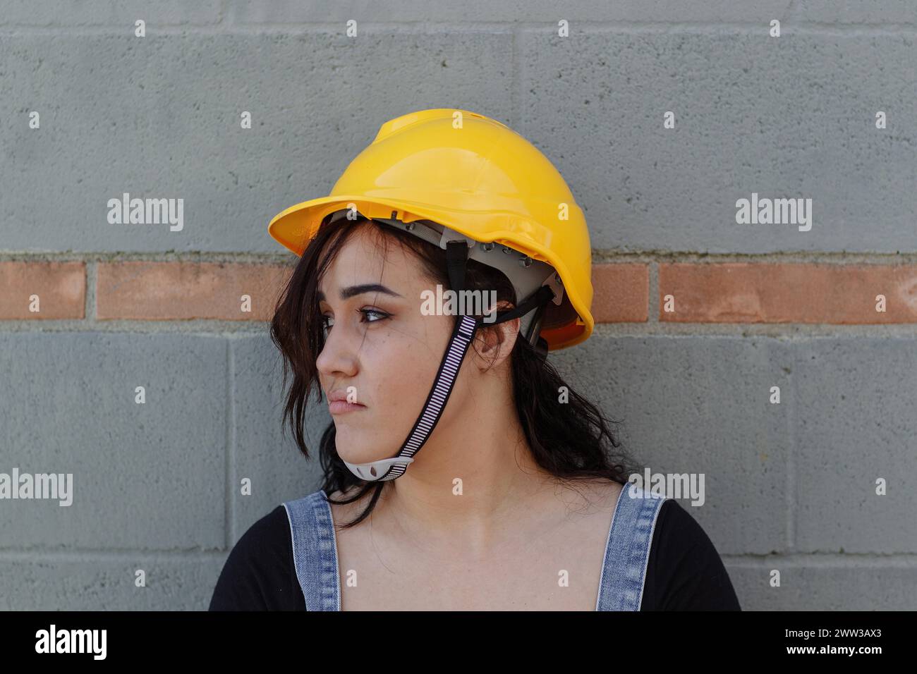 Working Woman in yellow hard hat loo king serious against a grey brick ...