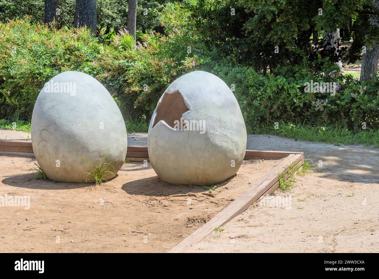 Two large egg sculptures, one cracked open, in a sandy park area, in ...